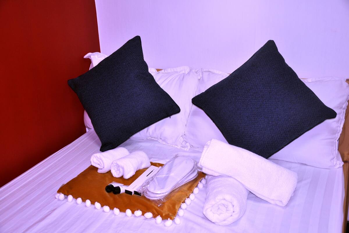 Cama bem arrumada com lençóis brancos, duas almofadas escuras e uma almofada decorativa marrom, além de um conjunto de toalhas brancas.