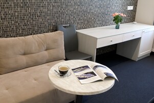 Quarto de hotel com sofá bege, uma mesa redonda pequena com uma chávena de café e uma revista, uma secretária com um vaso de flores e uma parede com azulejos.