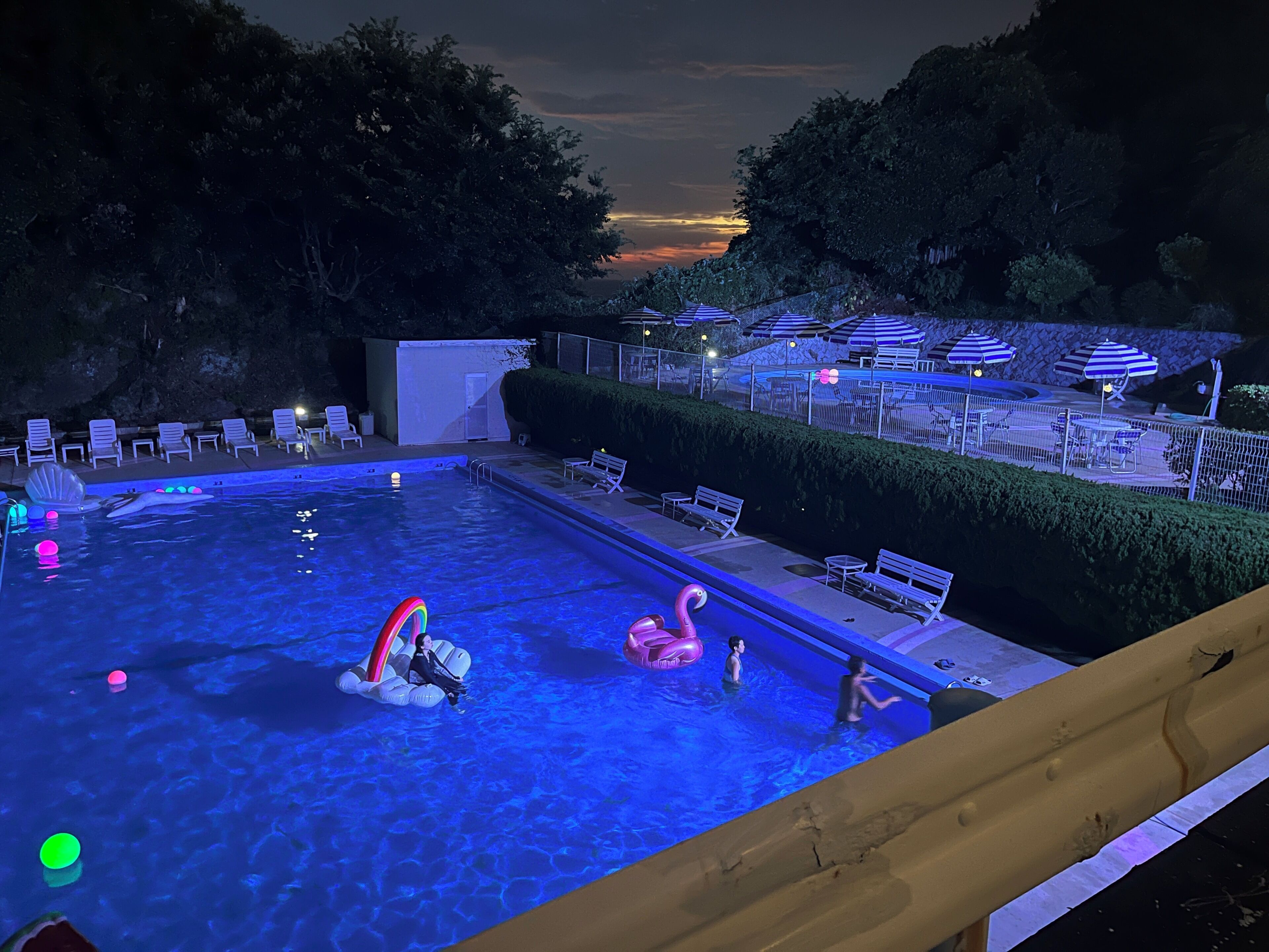 Uma piscina com espreguiçadeiras e guarda-sóis, cercada por árvores e uma vista do pôr do sol.