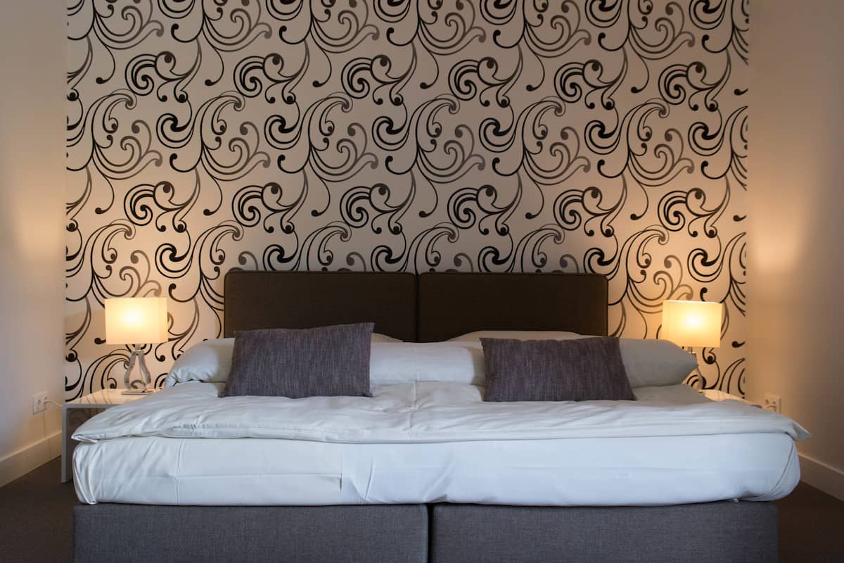 Uma cama de casal com cabeceira escura, duas luminárias de cabeceira e papel de parede estampado.