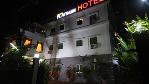 Um hotel chamado "Royal Platinum Hotel", com fachada moderna, várias janelas e plantas em vasos na área de entrada.