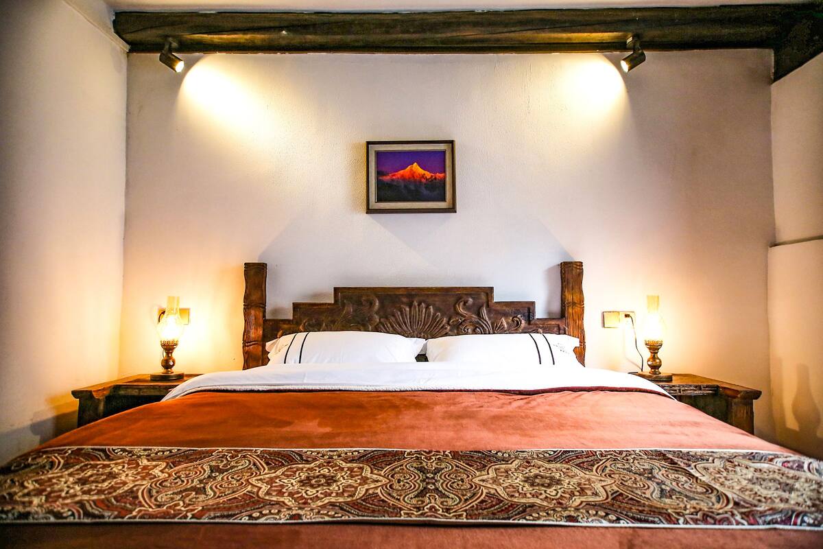 Uma cama com colcha estampada, duas luminárias de cabeceira, um quadro na parede e uma cabeceira de madeira.