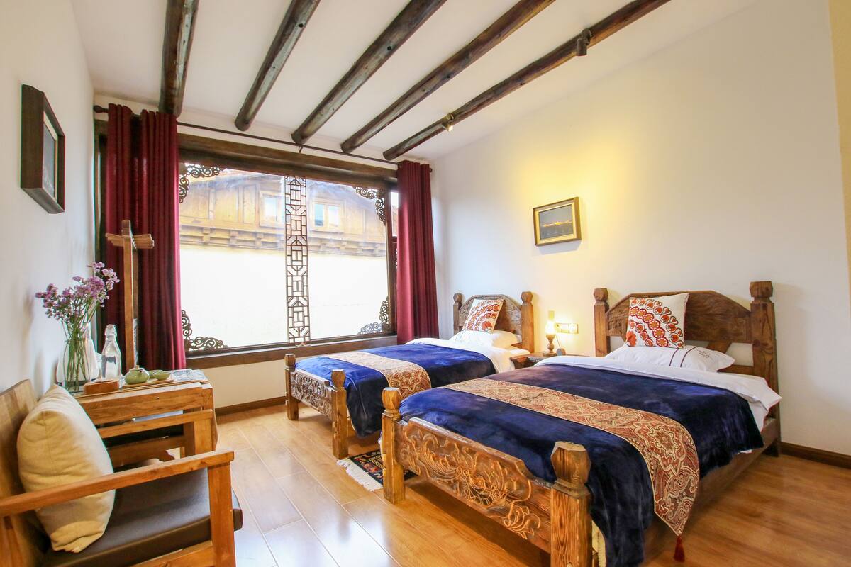 Quarto com duas camas de madeira, uma cadeira de madeira, uma mesinha com um vaso de flores e uma janela com cortinas.