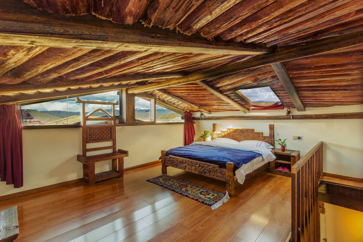 Quarto com teto de madeira, cama com colcha azul, cadeira de madeira, mesa de cabeceira com abajur e vista das montanhas pela janela.
