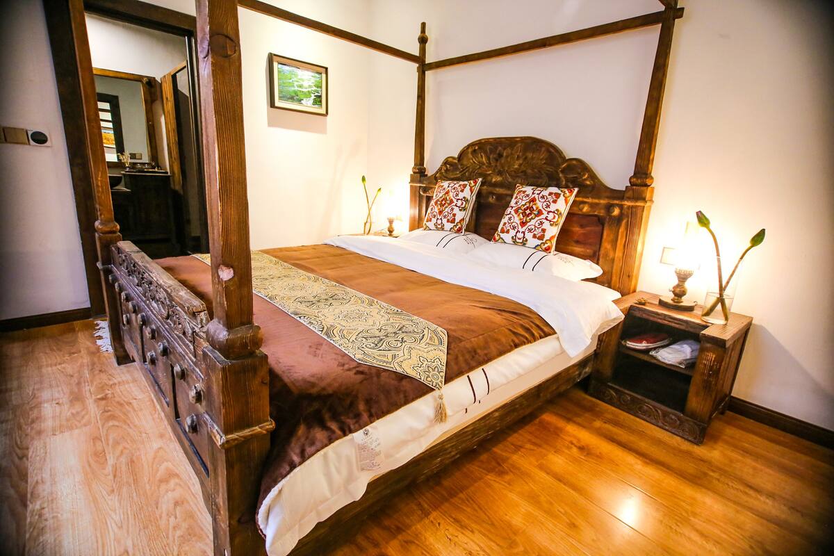 Uma cama com dossel, com cabeceira de madeira, mesa de cabeceira com abajur e espelho na parede.