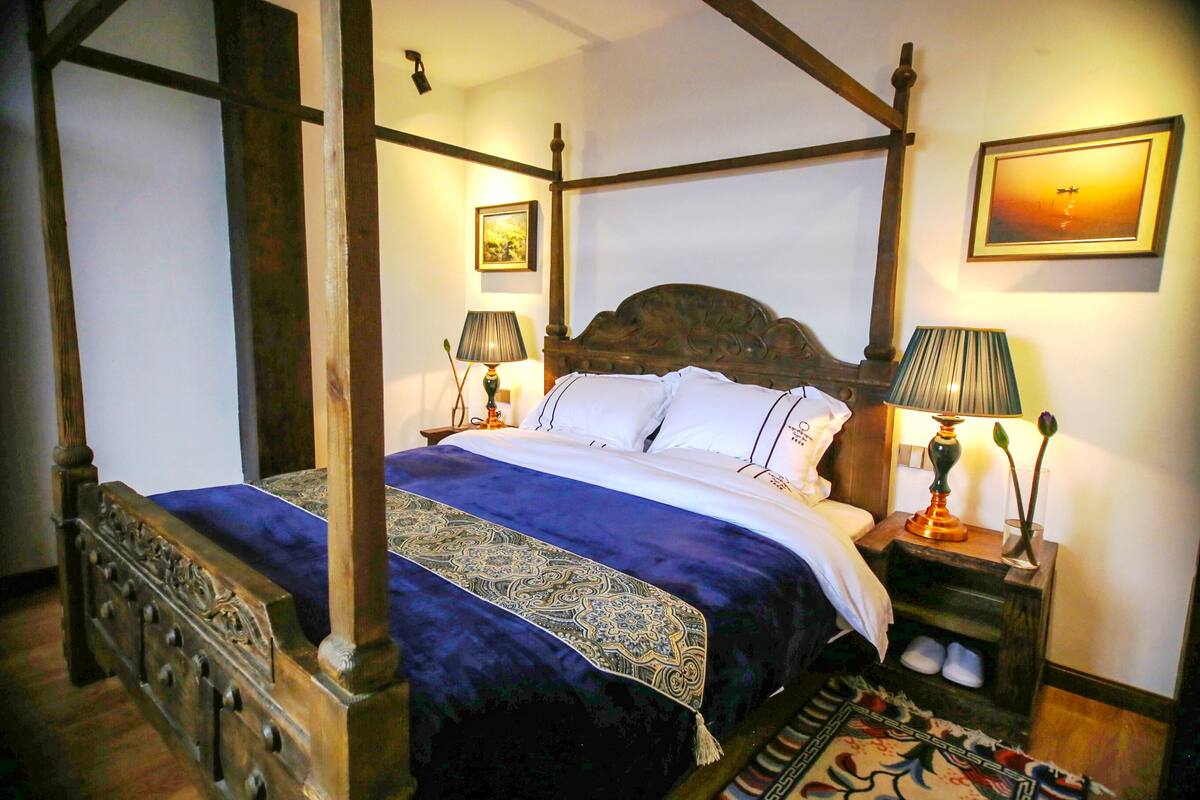 Uma cama de dossel com colcha azul e cabeceira decorativa. Há duas mesas de cabeceira com abajures e um pequeno estante com sapatos.