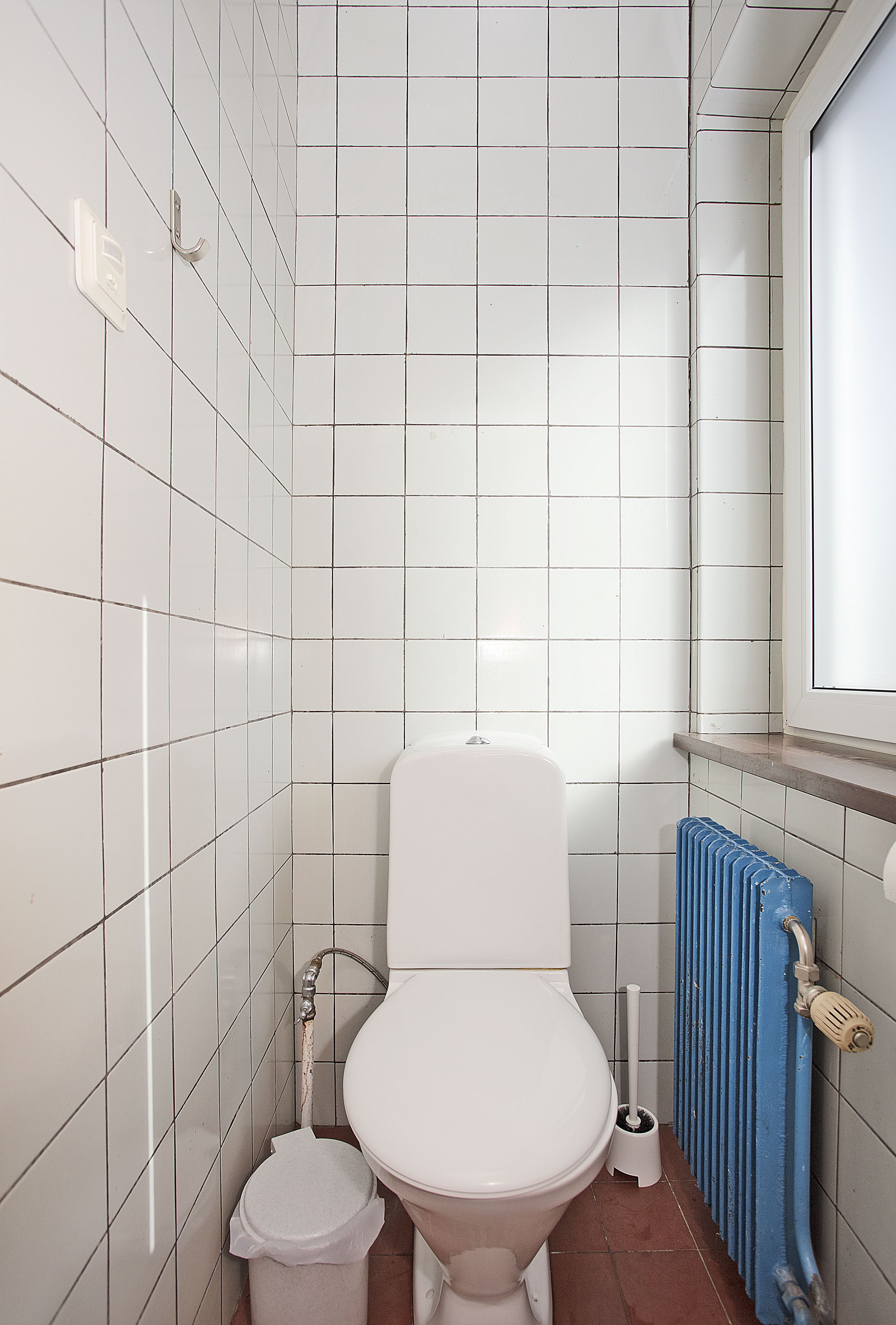 Um banheiro pequeno, com azulejos, um vaso sanitário branco, uma lixeira e um radiador azul.