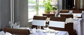 Restaurante com mesas preparadas para refeições, cadeiras com fitas brancas e plantas em vasos perto das janelas.