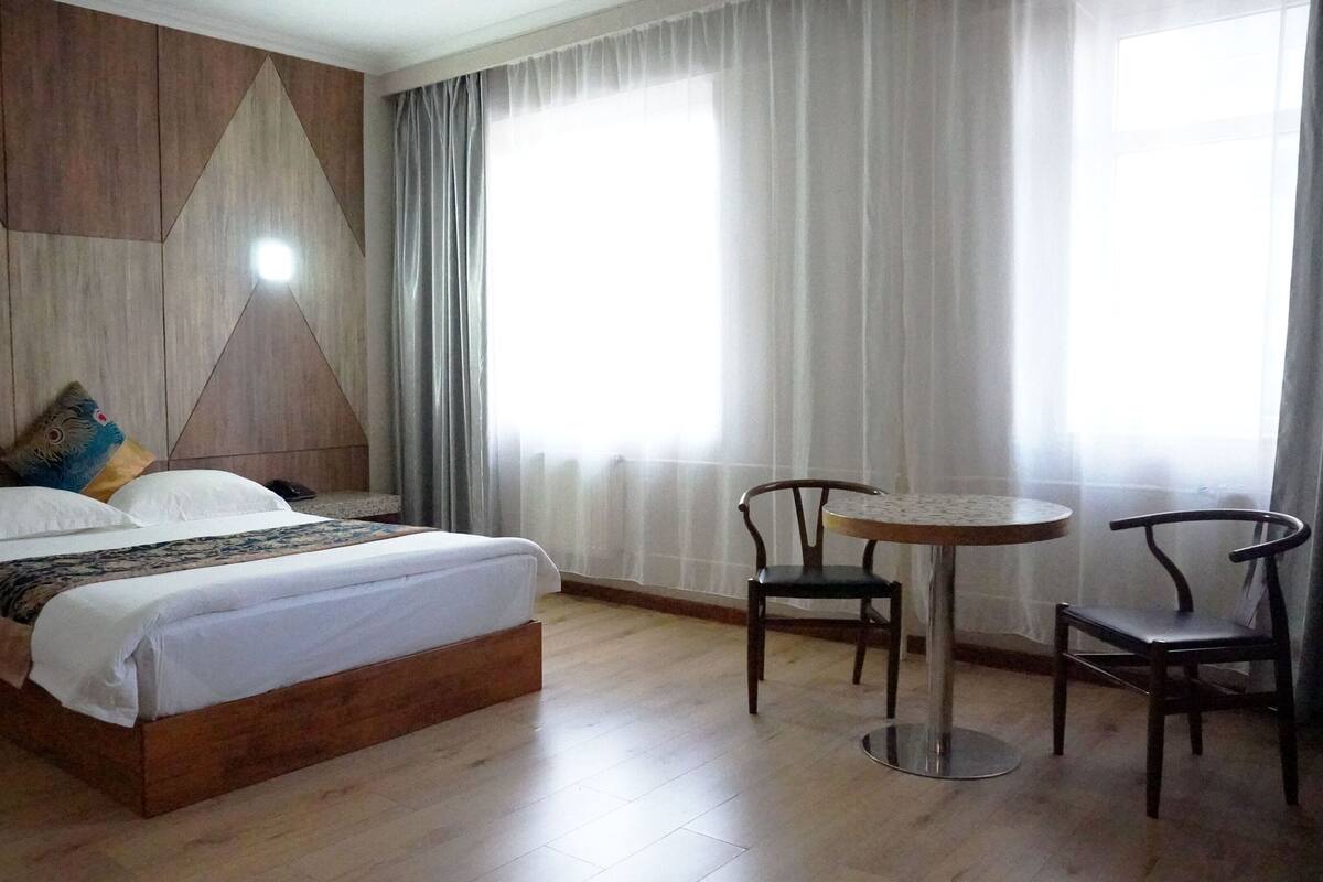 Quarto de hotel com cama, duas cadeiras, uma mesa pequena e uma janela grande com cortinas.