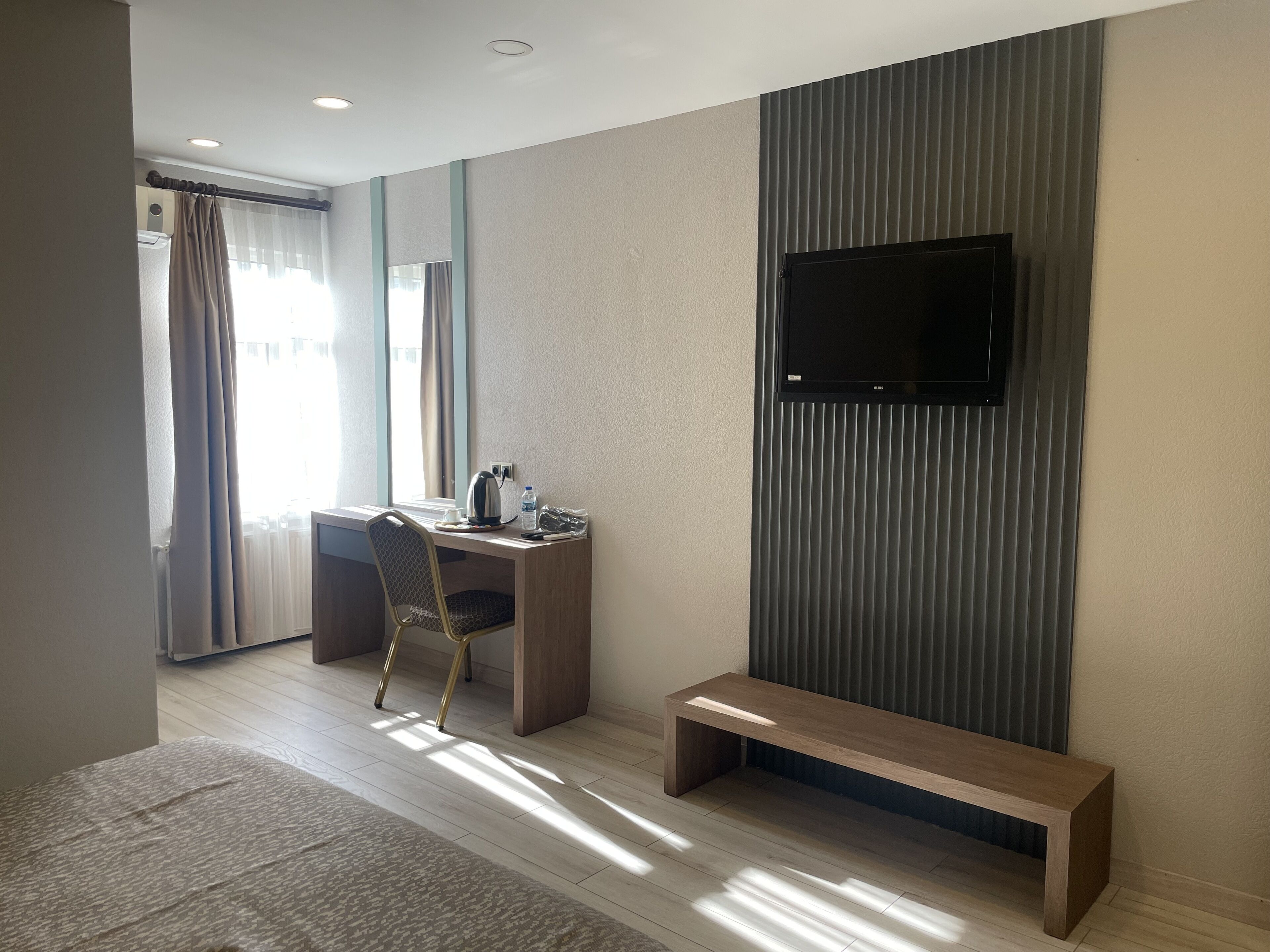 Quarto de hotel com escrivaninha, cadeira, banqueta e televisão na parede.