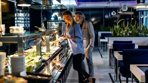 Uma área de buffet com diversos alimentos e algumas pessoas perto do balcão.