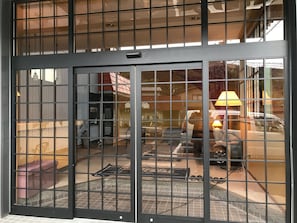 Visão através de um portão de metal com reflexos de carros estacionados e interior de um edifício.