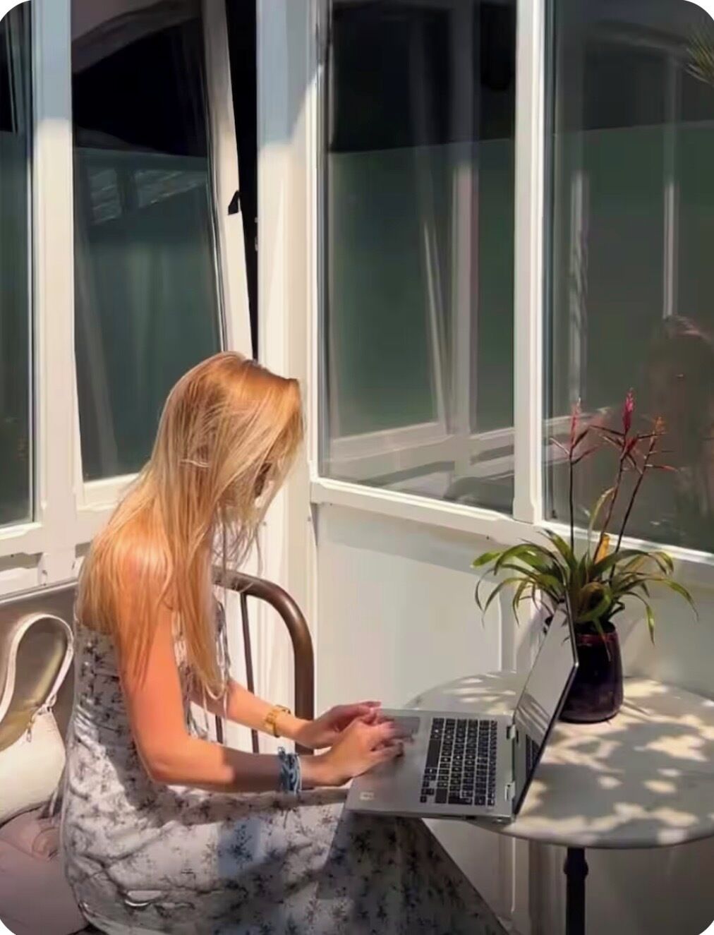 Uma pessoa sentada à mesa com um laptop, perto de uma janela com cortinas, e um vaso de planta sobre a mesa.