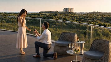 Um homem pedindo em casamento a uma mulher numa varanda com móveis de vime, uma mesa com garrafa e copos, e vista para o horizonte da cidade.