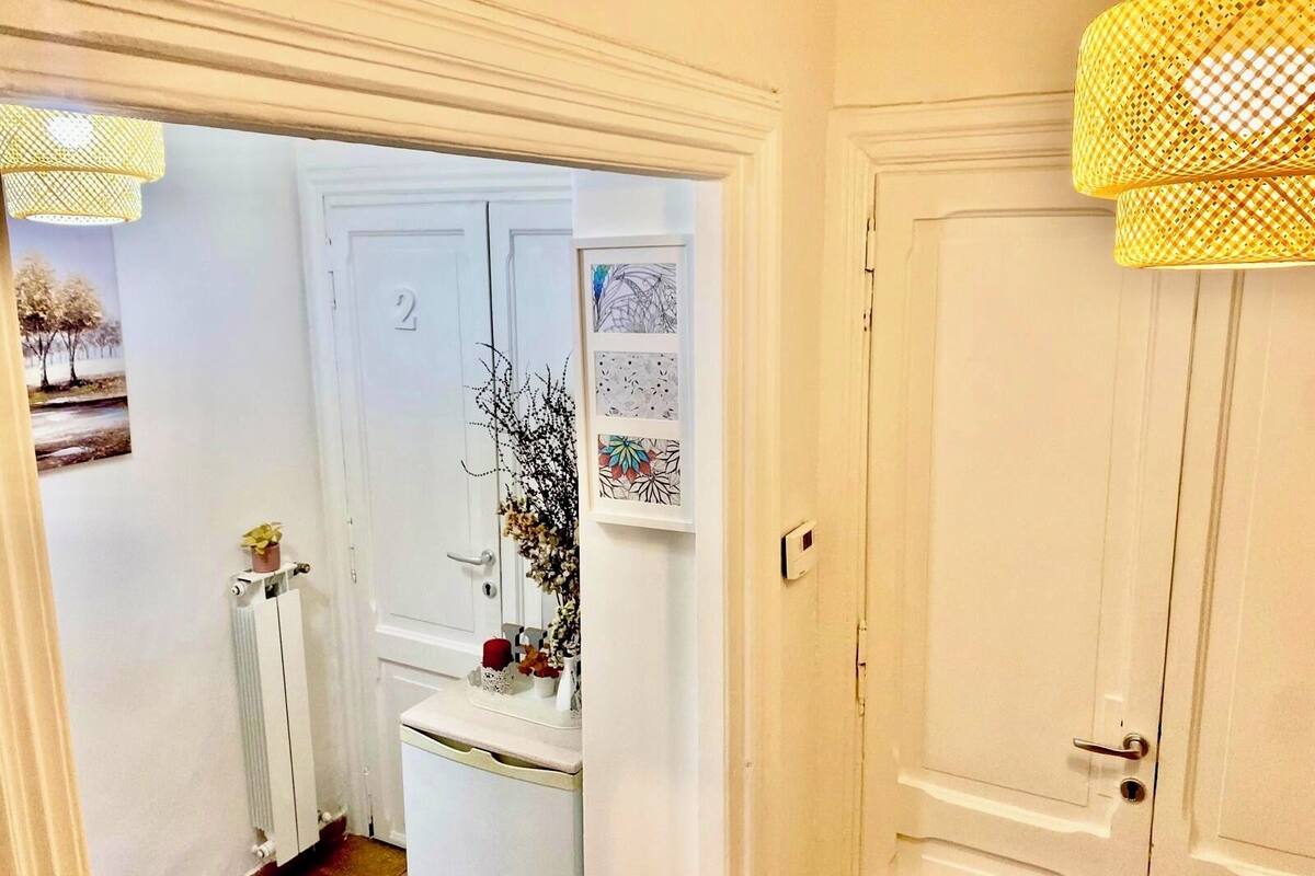Um corredor com uma porta branca marcada com o número 2, um radiador e uma pequena mesa com um vaso e uma vela vermelha.