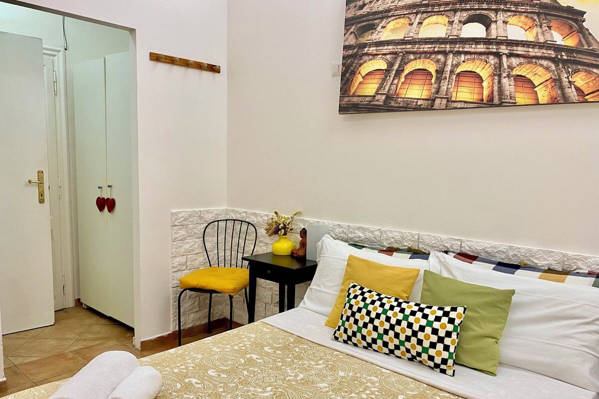 Quarto de hotel com uma cama, uma cadeira, um quadro do Coliseu e uma porta branca.
