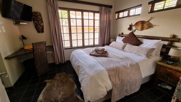 Quarto de hotel com cama, televisão, janela com cortinas, um objeto decorativo na parede e uma mesinha com abajur.