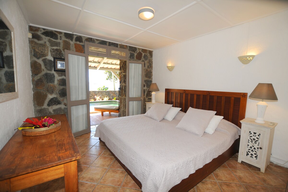 Quarto com parede de pedra, cama de madeira, mesa de cabeceira com abajur e cesta de vime sobre uma mesa de madeira.