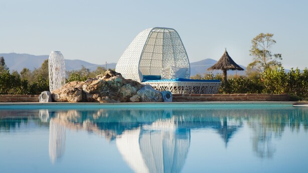Piscina com uma estrutura branca em forma de cúpula e um bangalô com telhado de palha ao fundo.