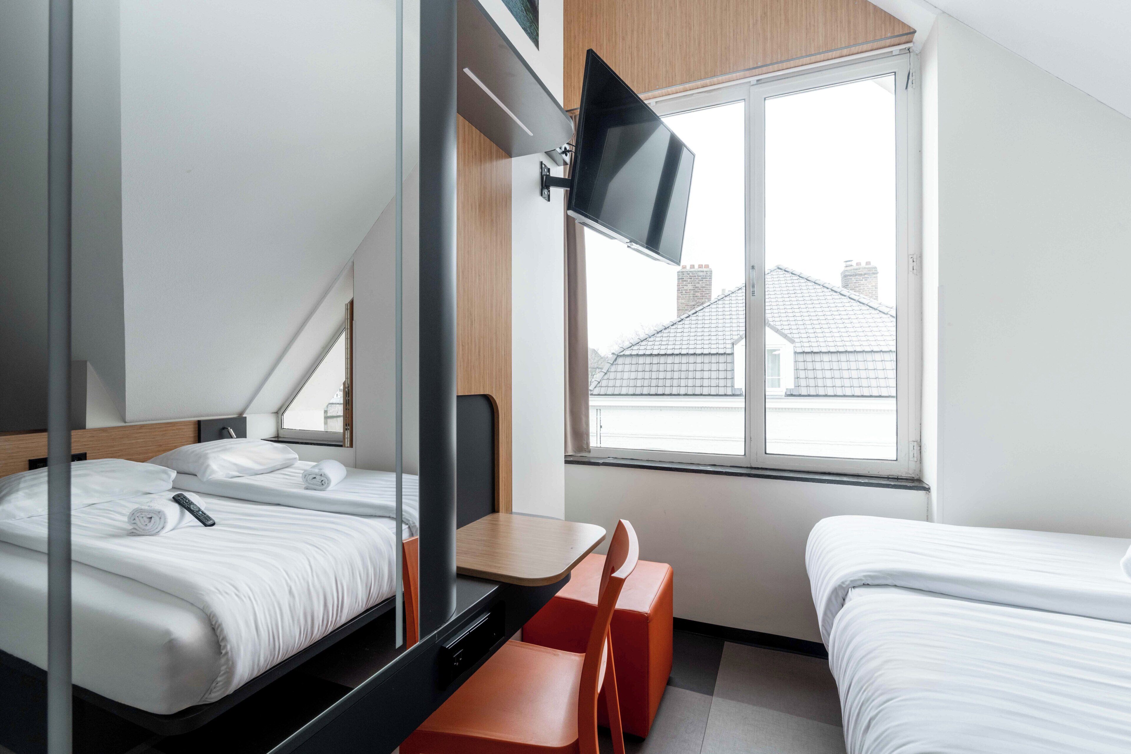 Quarto de hotel com cama beliche, escrivaninha, cadeira, televisão e janela com vista para prédios.