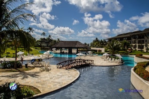 Resort com uma piscina grande, espreguiçadeiras, palmeiras e um pavilhão coberto.