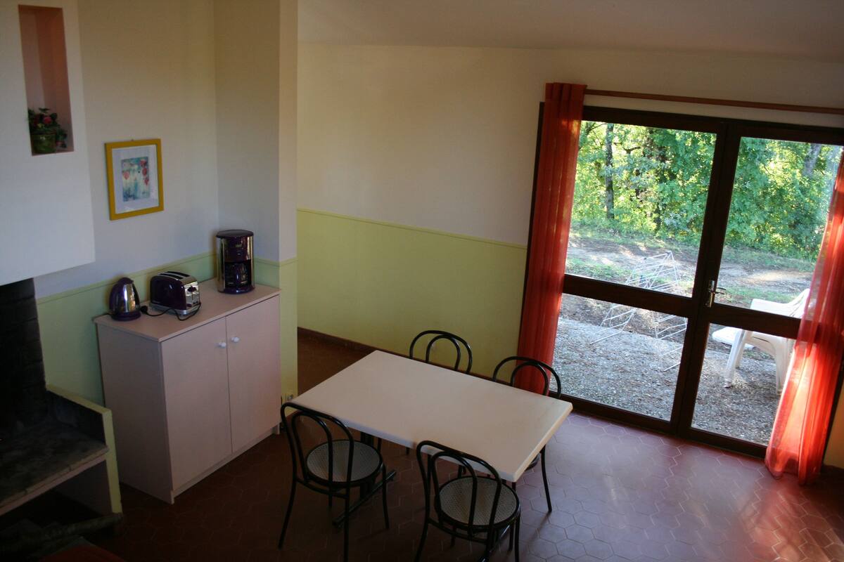 Quarto com uma mesa branca e quatro cadeiras, um armário com eletrodomésticos, um quadro na parede e uma janela grande com cortinas.