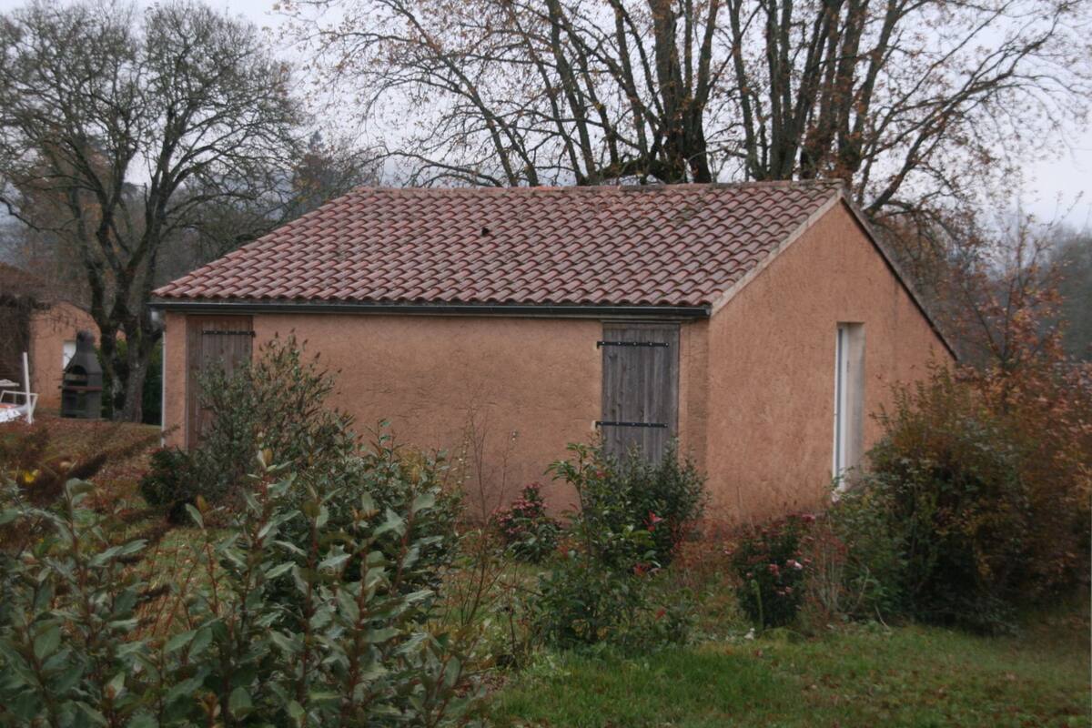 Um pequeno edifício térreo com telhado de telhas, rodeado por vegetação e árvores.