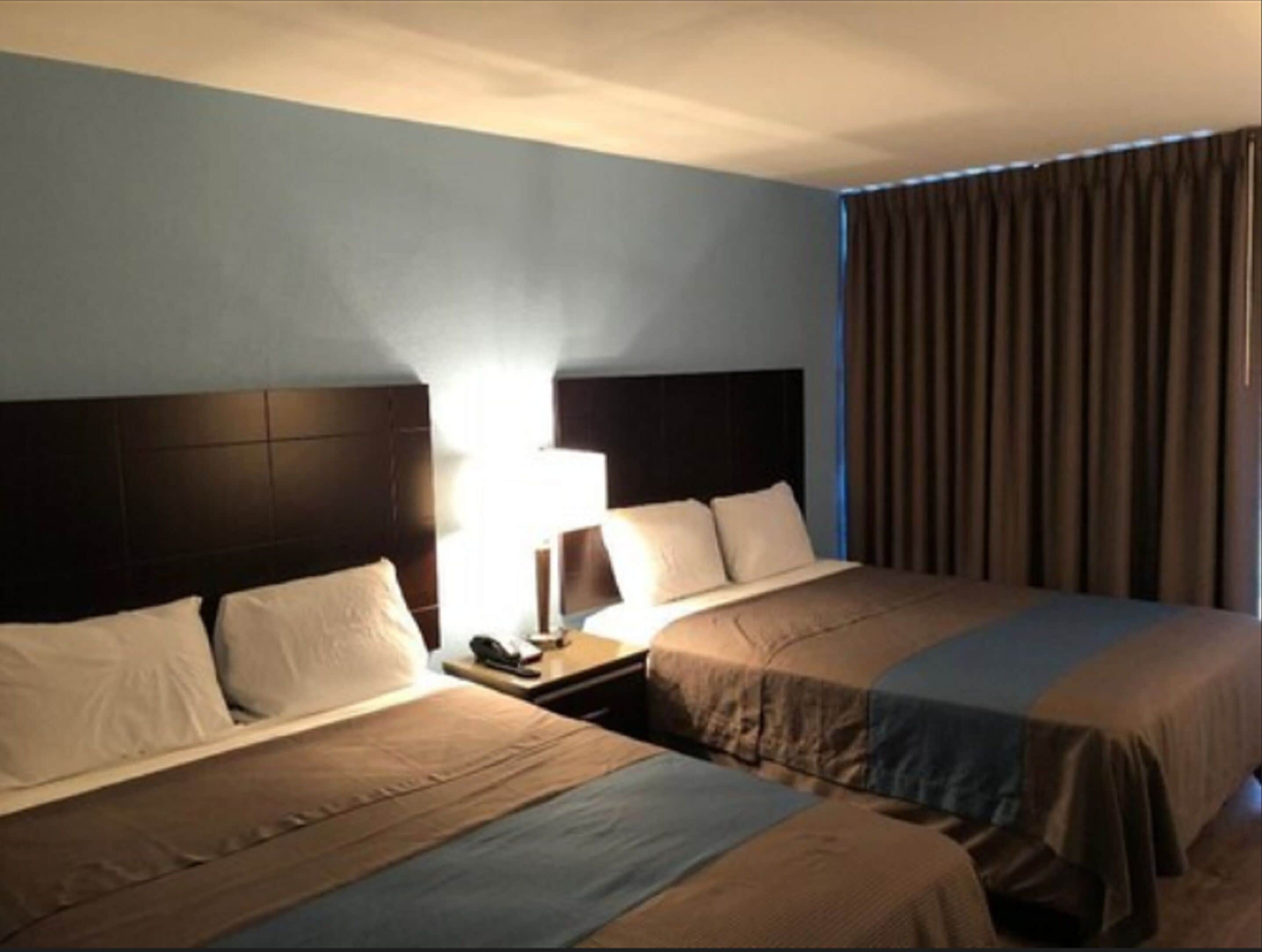 Quarto de hotel com duas camas, uma mesa de cabeceira, uma luminária e uma janela com cortinas.