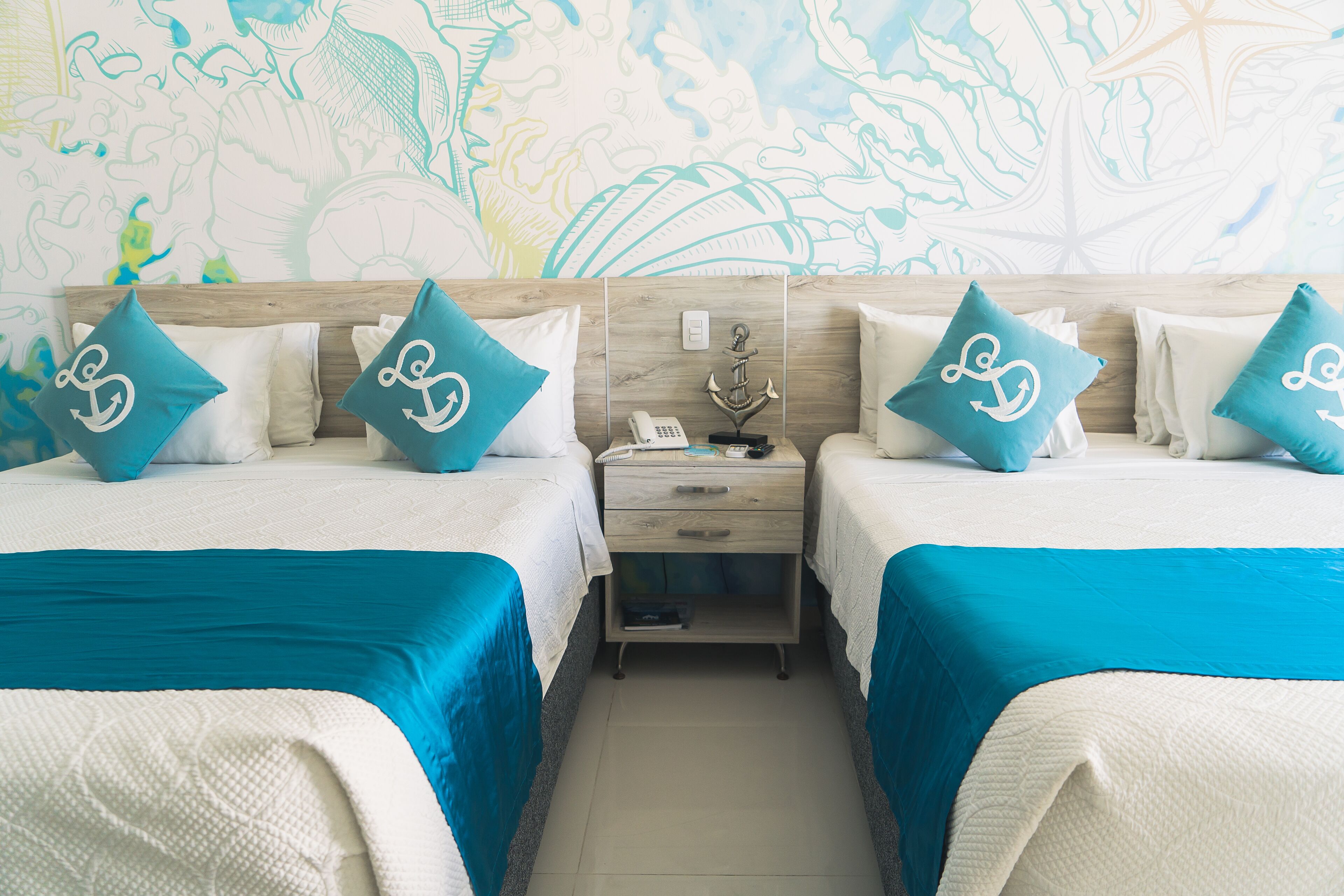 Quarto de hotel com duas camas, cada uma com colcha azul e almofadas a combinar. Há uma mesa de cabeceira entre as camas, com um telefone e um pequeno objeto decorativo.