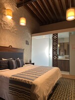 Quarto com cama, mesas de cabeceira e banheiro visível através de uma porta aberta.