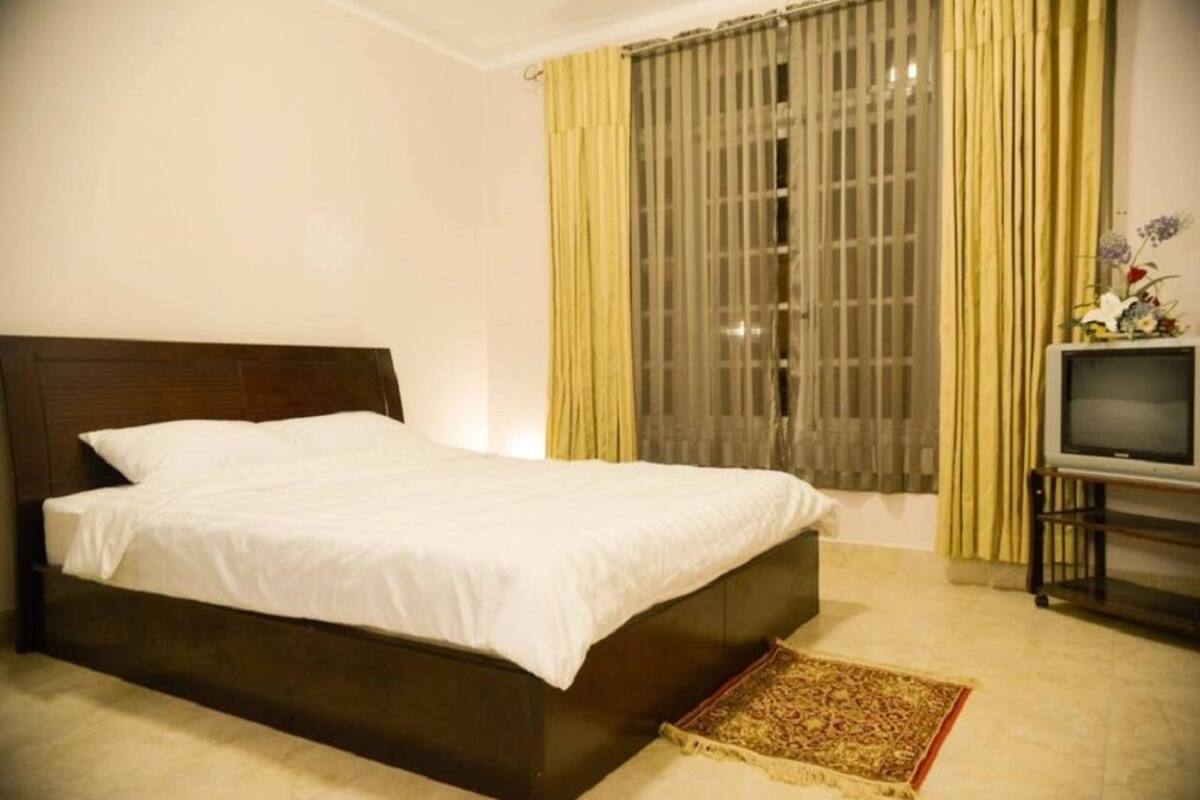 Cama bem arrumada com lençóis brancos num quarto com uma janela grande coberta por cortinas amarelas.