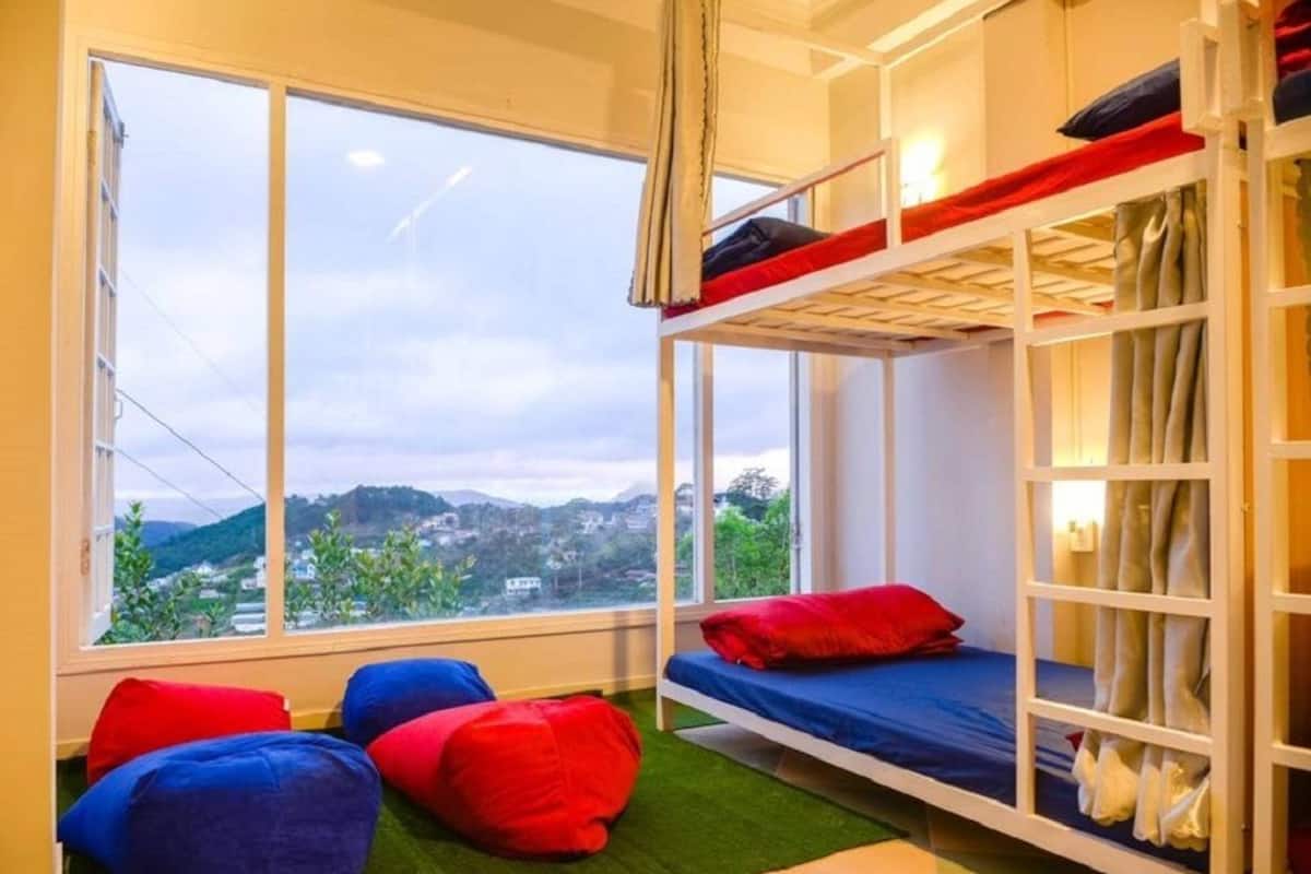 Quarto com beliche e vista para uma cidade e colinas. Há sacos de feijão coloridos e uma pequena mesa.