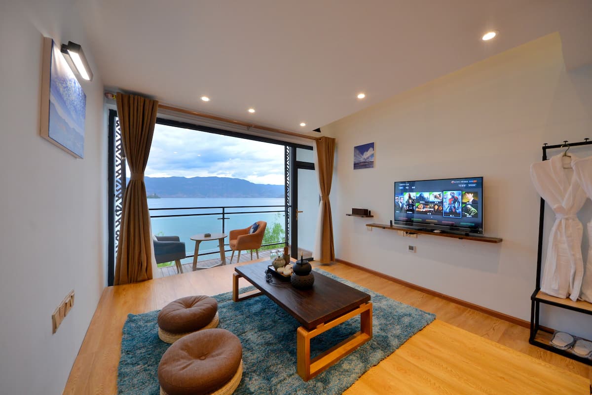 Uma sala de estar moderna com uma janela grande, uma televisão de tela plana, uma mesa de centro de madeira e um sofá com almofadas.