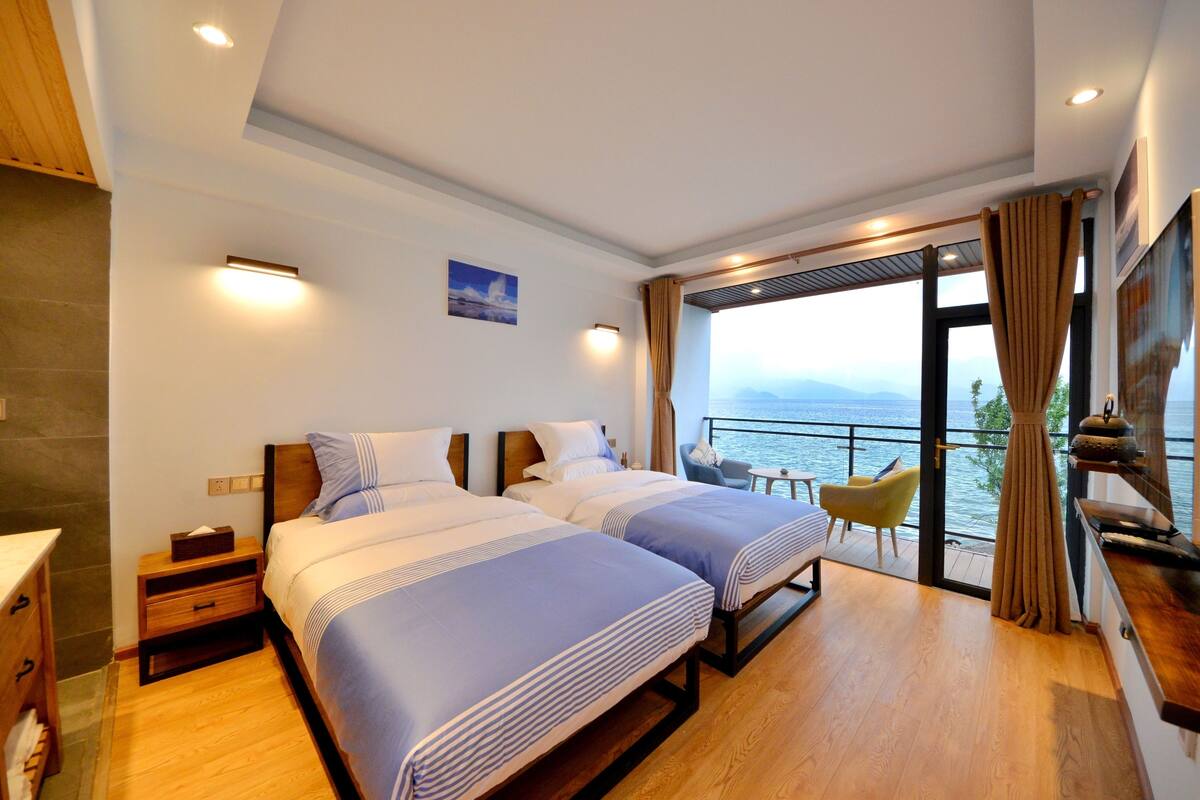Quarto de hotel moderno com duas camas, piso de madeira, varanda com vista e um pequeno espaço de estar.