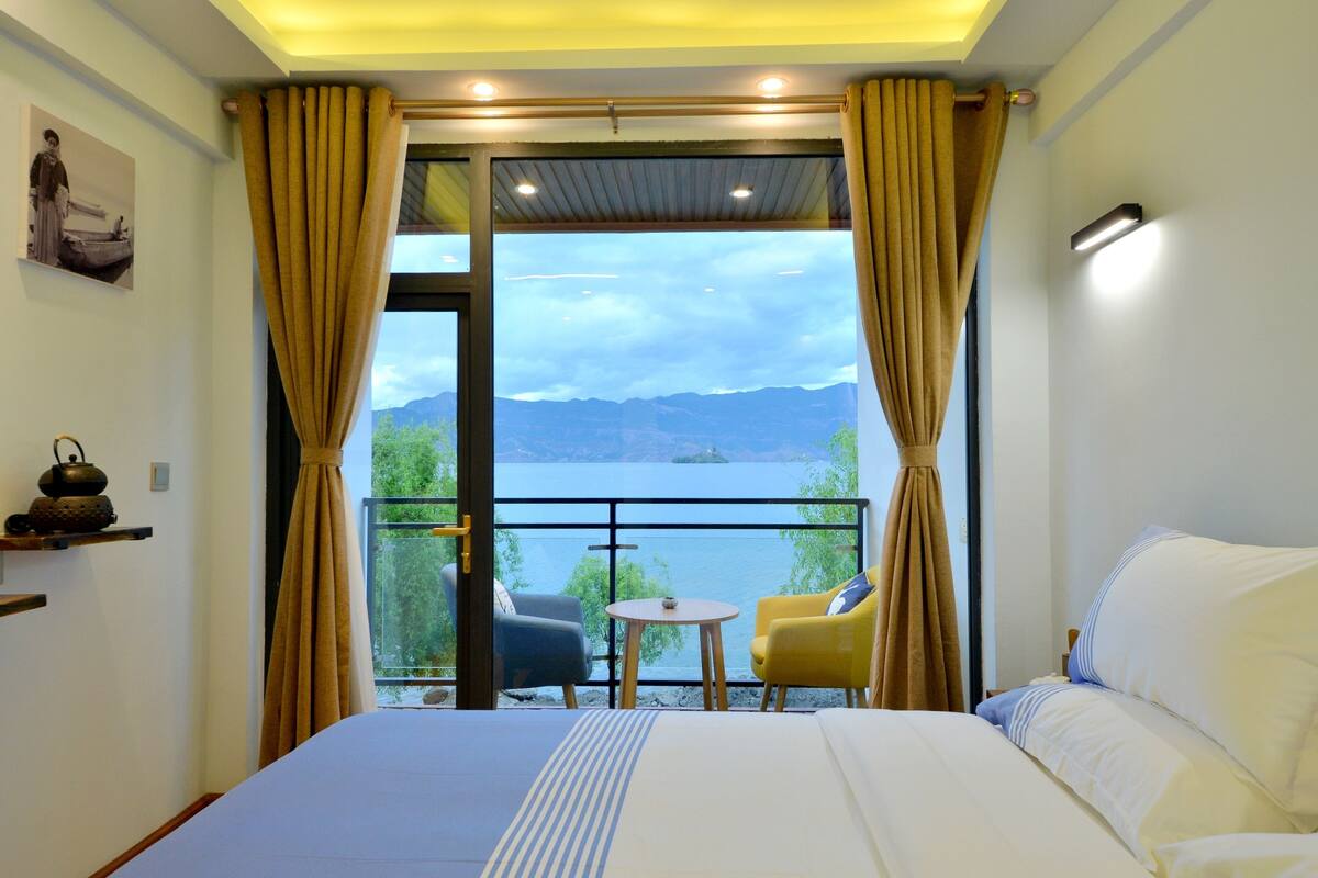 Quarto de hotel com cama, uma mesinha, duas cadeiras e vista para um lago e montanhas.