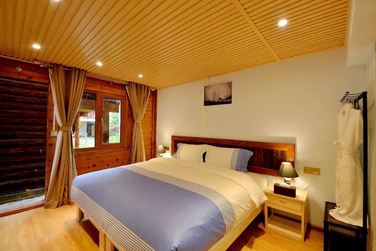 Quarto de cabana de madeira com uma cama grande, uma mesa de cabeceira, uma luminária e uma janela com cortinas.