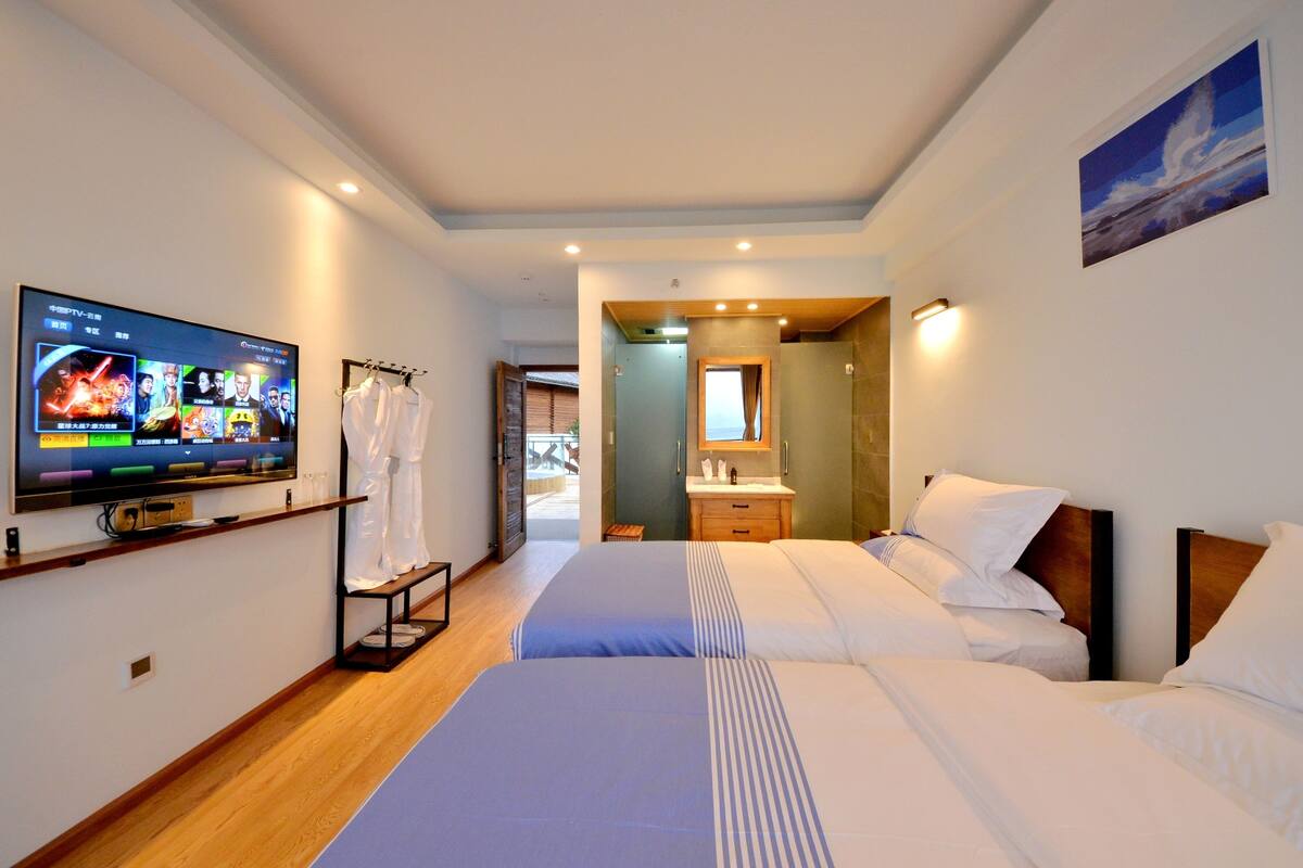 Quarto de hotel com duas camas, televisão na parede, casa de banho com espelho e um quadro na parede.