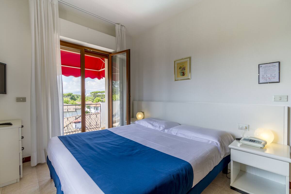 Quarto de hotel com cama, mesa de cabeceira e vista através de uma porta de vidro.