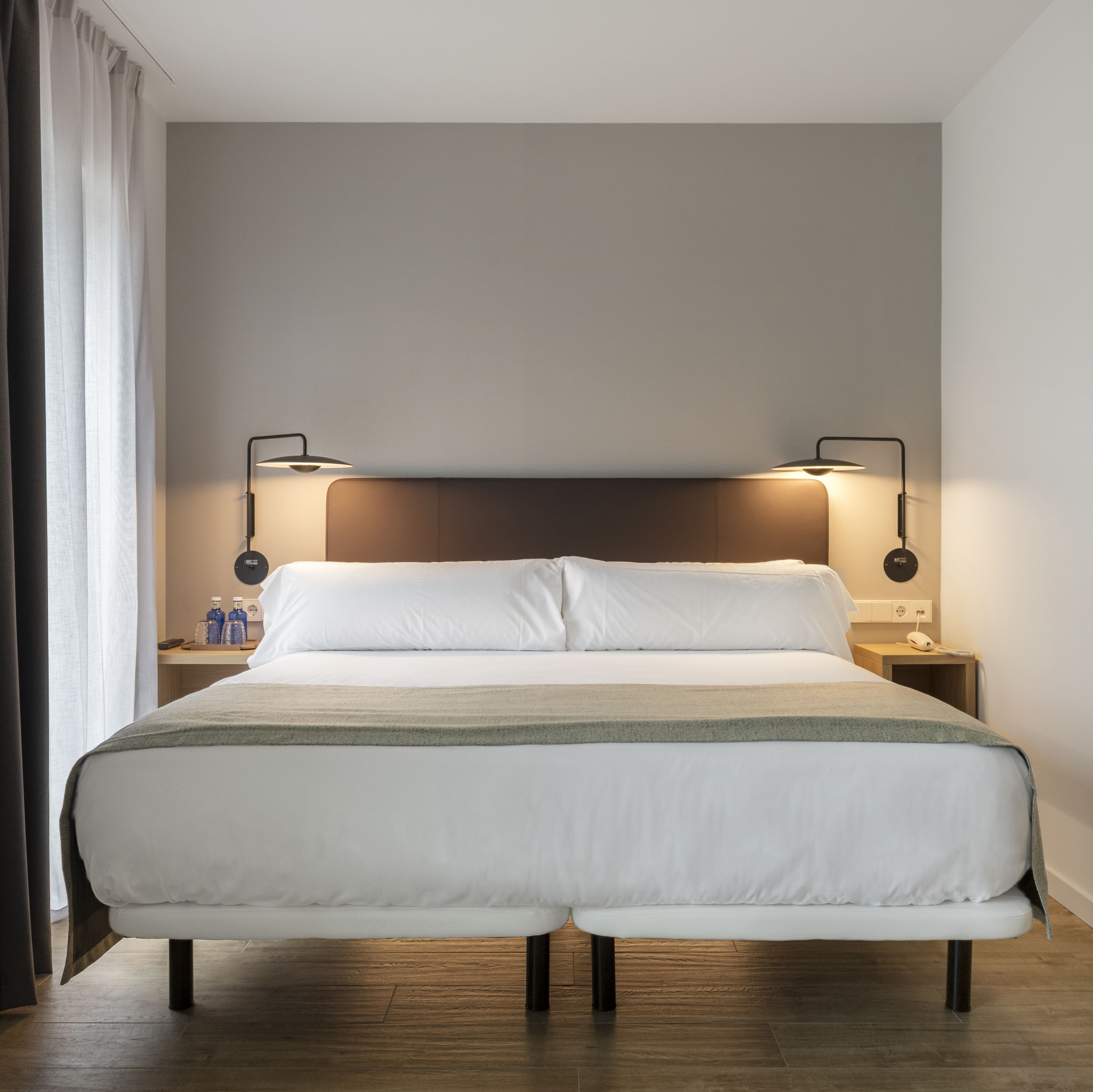 Uma cama de casal bem arrumada com um cobertor cinza, duas mesas de cabeceira com abajures e piso de madeira.