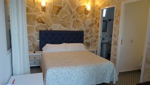 Quarto com parede de pedra, cama com cabeceira escura, mesas de cabeceira e banheiro visível por uma porta aberta.