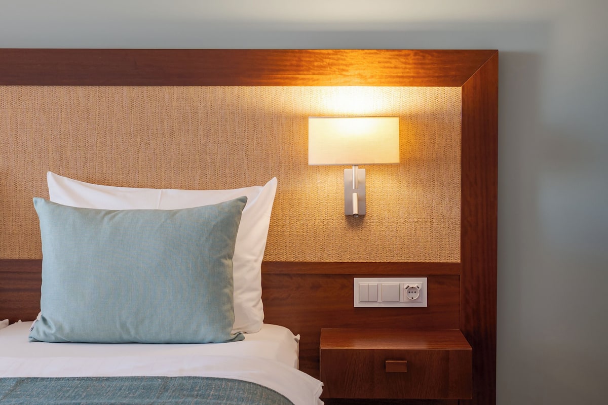 Quarto de hotel com uma cama grande, uma mesa de cabeceira com abajur e cabeceira de madeira.