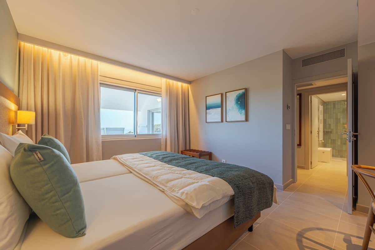 Quarto de hotel com uma cama grande, uma mesa de cabeceira, uma luminária e vista para o exterior.