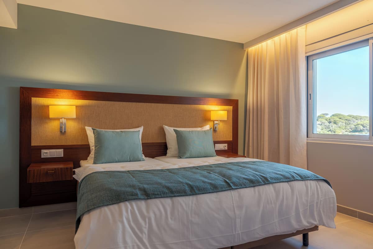 Quarto de hotel com uma cama grande, duas luminárias de cabeceira, uma mesa de apoio e uma janela com cortinas.