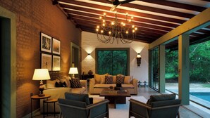 Uma sala de estar moderna com um sofá, duas poltronas, uma mesa de centro e um candelabro. Há quadros nas paredes e uma janela com vista para o verde.