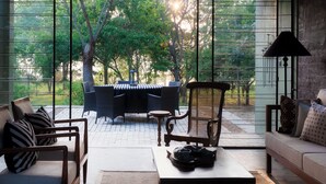 Uma sala de estar espaçosa com vista para um jardim através de grandes janelas. Há um sofá, uma poltrona e uma pequena mesa com um cesto.