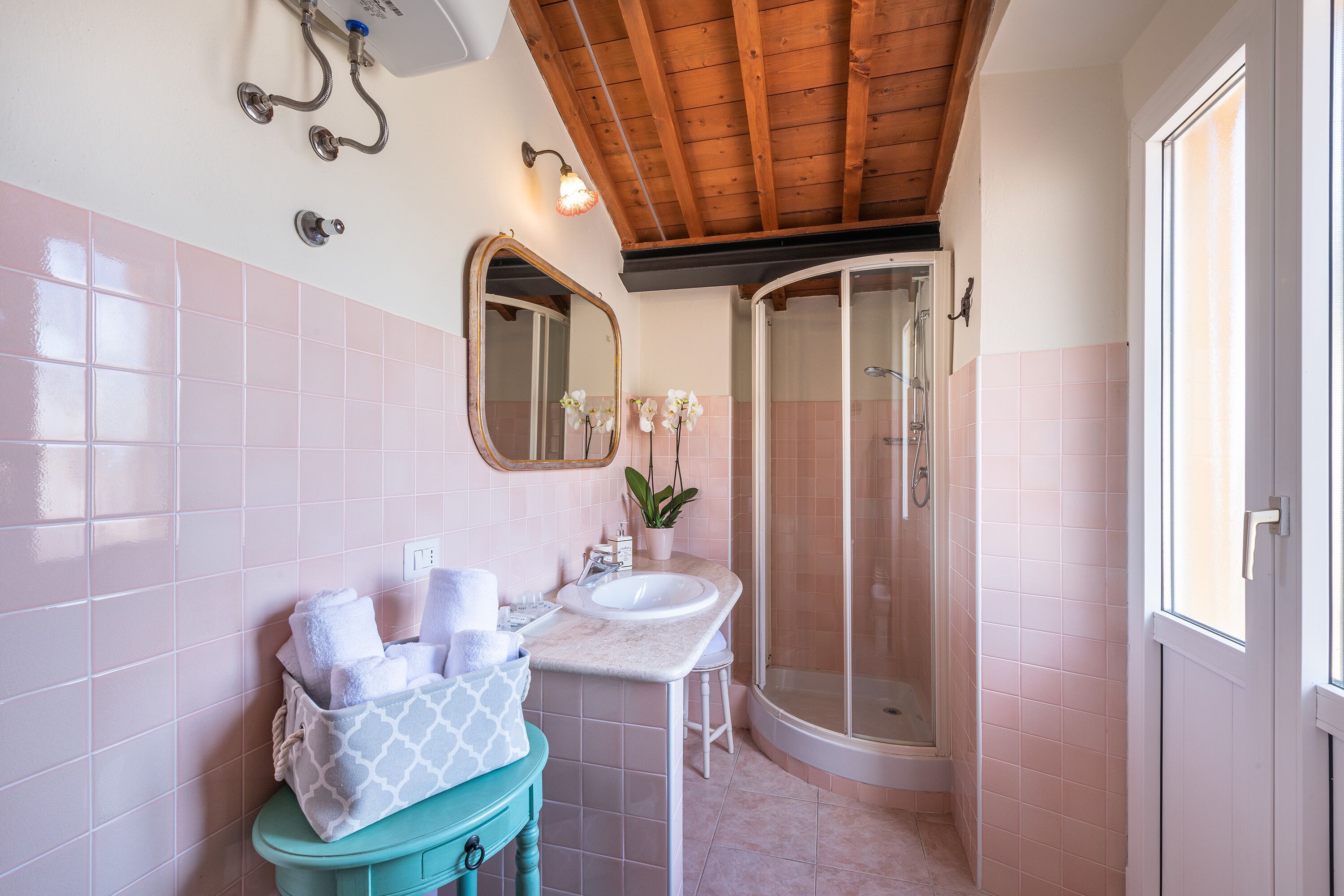 Casa de banho com azulejos cor-de-rosa, uma pia branca, um espelho, um chuveiro e um toucador verde-água.