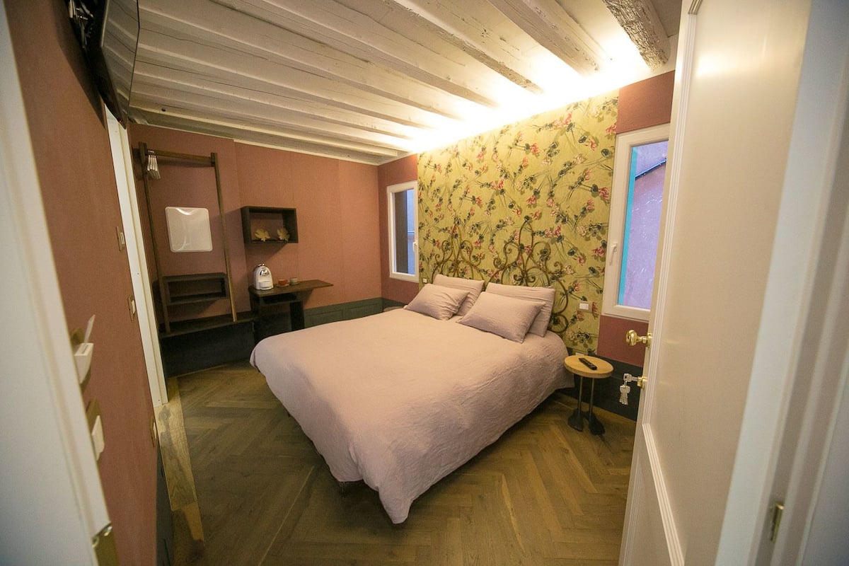 Quarto com uma cama grande, uma mesa de cabeceira, uma pequena mesa, televisão na parede e janela com vista.