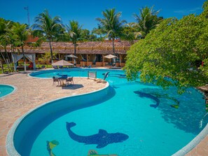 Piscina com mural de golfinho, rodeada por palmeiras e um deck revestido com azulejos.