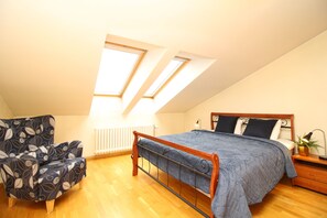 Um quarto com teto inclinado, uma cama com colcha azul, uma cadeira com padrão de folha e uma mesa de cabeceira com abajur.