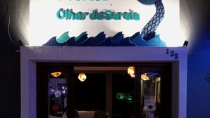 Entrada de um prédio com uma placa indicando "Pousada Olhar da Sereia" e uma decoração de rabo de sereia sobre a entrada.
